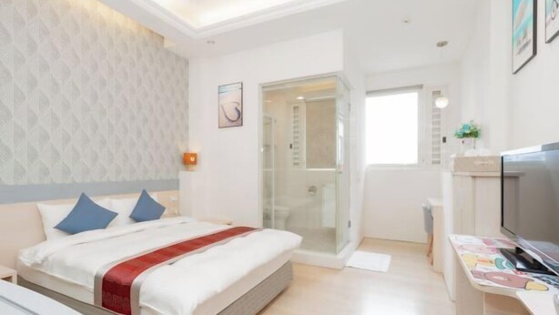 Quarto de hotel moderno com cama, escrivaninha com computador, TV e área de chuveiro envidraçada.