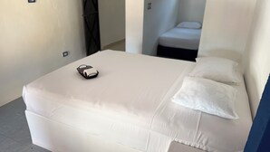 Uma cama bem arrumada com colchão e almofadas brancas, uma pequena cesta preta com um objeto branco e uma parede lisa com uma porta ao fundo.