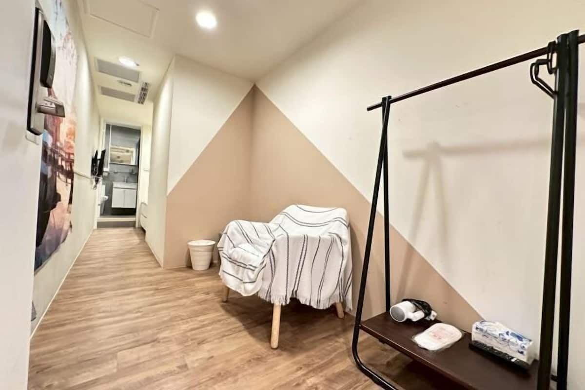 Um corredor com piso de madeira, uma cadeira coberta, uma lixeira e uma prateleira com objetos.