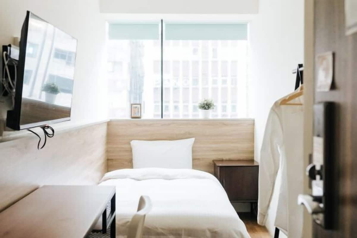 Quarto de hotel com cama, televisão, janela com persianas, mesa de cabeceira e guarda-roupa.