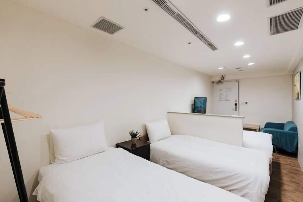 Quarto de hotel com duas camas, uma escrivaninha, uma cadeira e uma pequena mesa com uma planta.