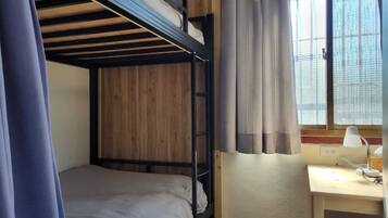 Uma cama beliche com cabeceira de madeira, uma escrivaninha com abajur e uma janela com cortinas.