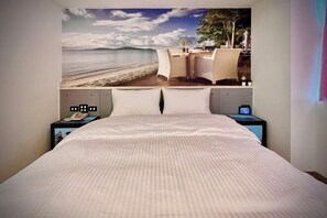 Um quarto com cama, mesa de cabeceira, televisão na parede e arte decorativa com tema de praia.