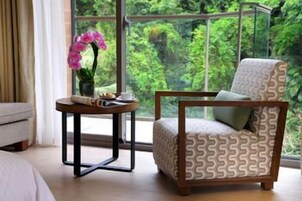 Um quarto com janela, uma cadeira, uma mesinha e um vaso com flores.