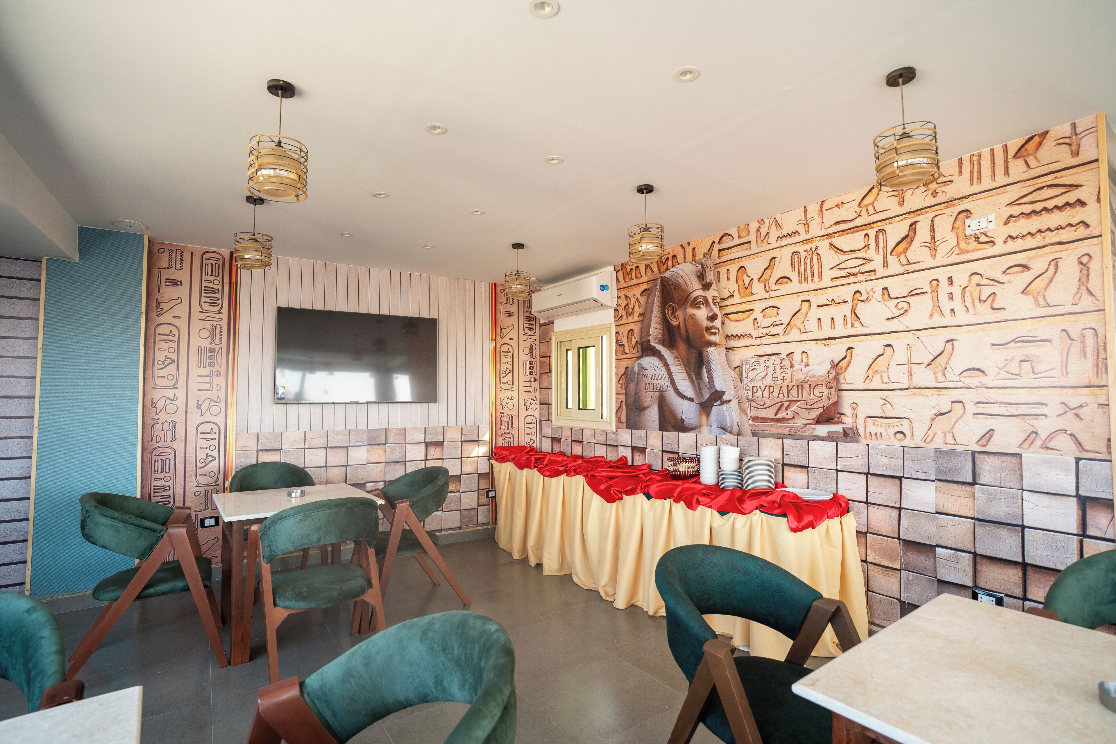 Interior de restaurante com cadeiras estofadas em verde, uma mesa comprida com toalha vermelha e paredes decoradas com hieróglifos.