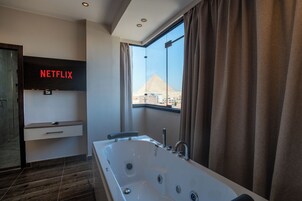 Uma casa de banho moderna com uma banheira grande, uma televisão embutida a exibir 'NETFLIX' e vista para uma pirâmide através de uma janela.