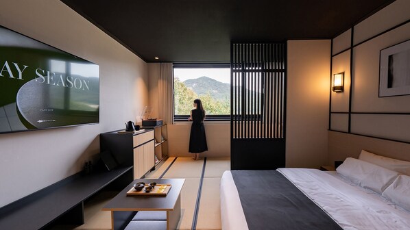 Quarto de hotel moderno com uma televisão grande exibindo 'PLAY LIST', uma cama, uma escrivaninha com um laptop, uma pequena mesa com tigelas e vista para montanhas pela janela.