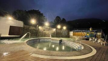 Área de piscina aquecida ao ar livre com deck de madeira, rodeada por paredes de pedra e iluminada por luzes.