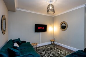 Uma sala de estar com um sofá verde, uma televisão de tela plana na parede, um abajur e um tapete estampado.