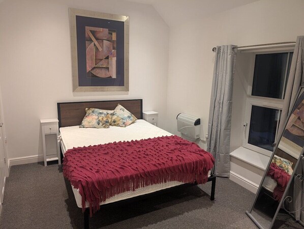 Um quarto com cama, mesa de cabeceira, janela com cortinas e uma obra de arte pendurada na parede.