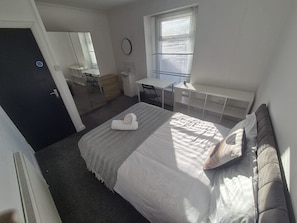 Quarto com cama, escrivaninha, cadeira, espelho e janela.