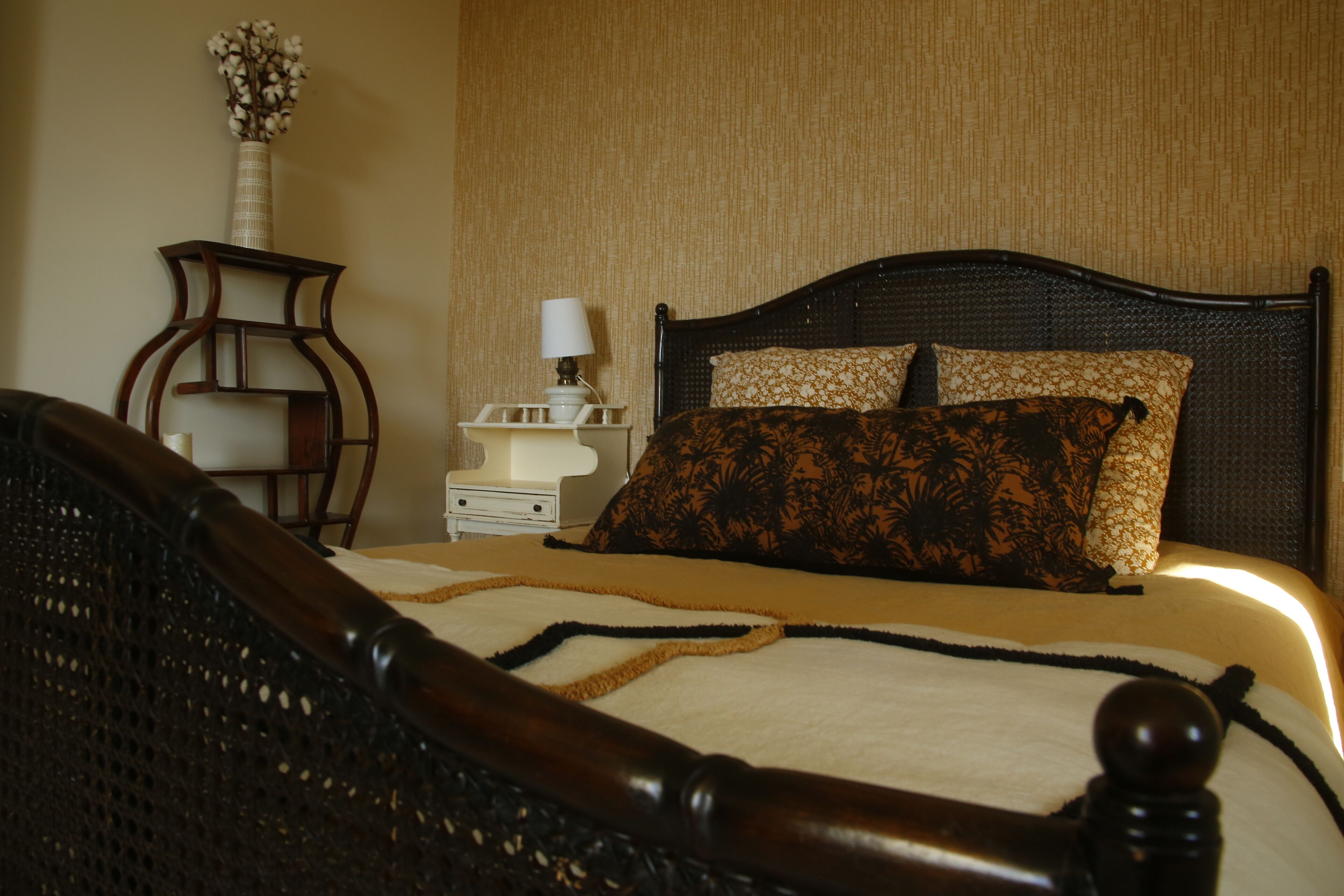 Uma cama bem arrumada com cabeceira escura, mesa de cabeceira com abajur e um vaso decorativo em cima.