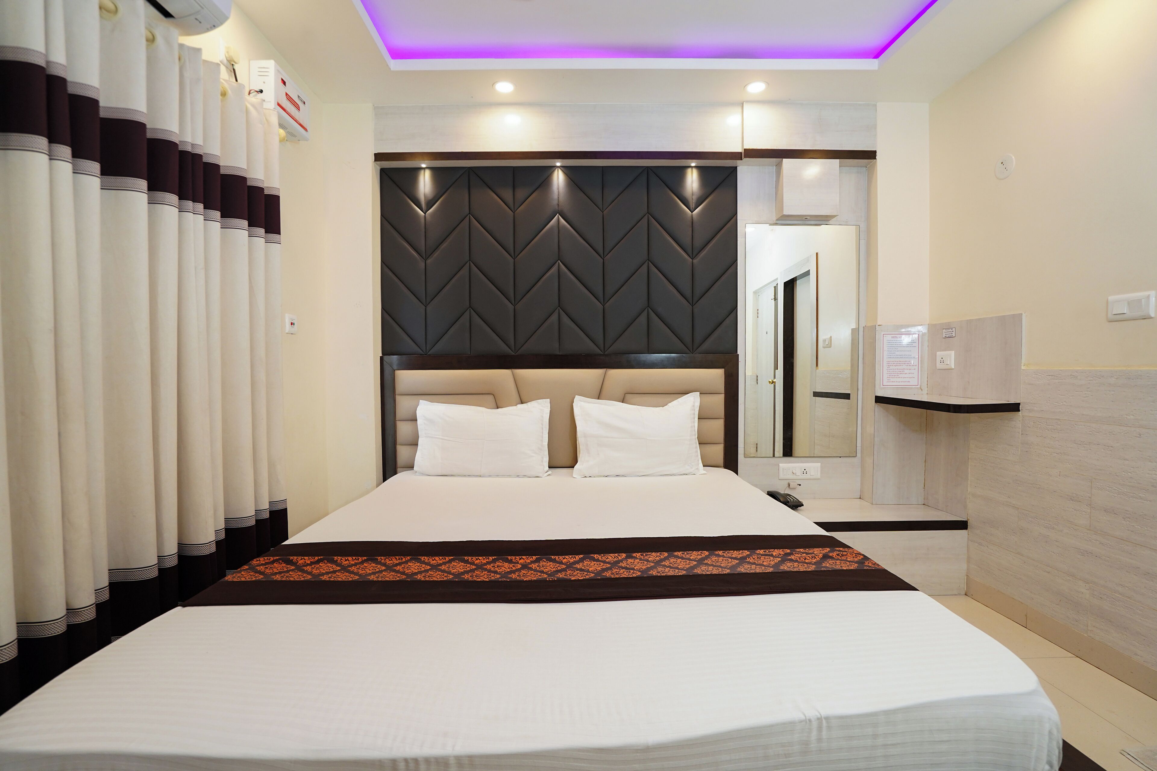 Quarto de hotel com uma cama grande, cabeceira com padrão em ziguezague, espelho e um espaço pequeno para o banheiro.