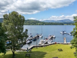 Uma marina com barcos atracados, uma área gramada com bancos e vista para colinas ao fundo.