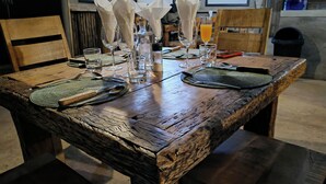 Mesa de madeira rústica posta com copos, guardanapos e talheres, indicando uma área de refeições acolhedora, possivelmente histórica.