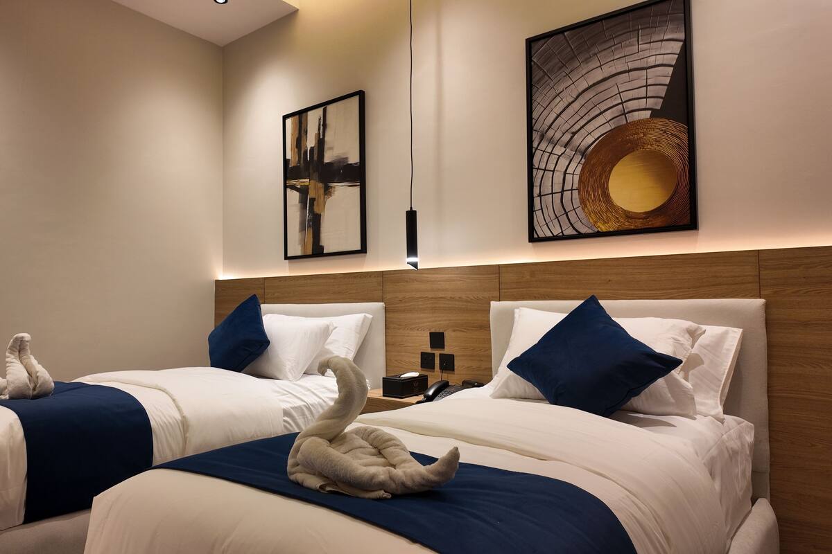 Quarto de hotel com duas camas, cada uma com colcha azul e branca, cabeceira de madeira e uma obra de arte pendurada na parede.