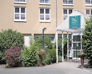 Entrada de hotel com placa indicando Quality Hotel Erlangen, rodeada por vegetação e uma galeria coberta.