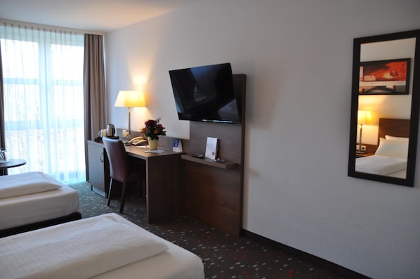 Quarto de hotel com cama, escrivaninha com cadeira, televisão na parede e janela com cortinas.