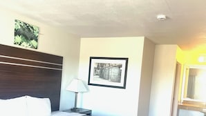 Quarto de hotel com cama, mesa de cabeceira, luminária, quadro na parede e banheiro visível por uma porta aberta.