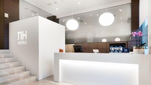 Uma área de recepção de hotel moderna, com um balcão branco, um visor digital e uma escadaria que dá acesso aos andares superiores.