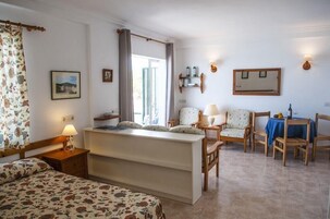 Quarto de hotel acolhedor com cama, escrivaninha, cadeira e uma pequena mesa. Há uma janela com cortinas, uma prateleira com garrafas e um quadro emoldurado.