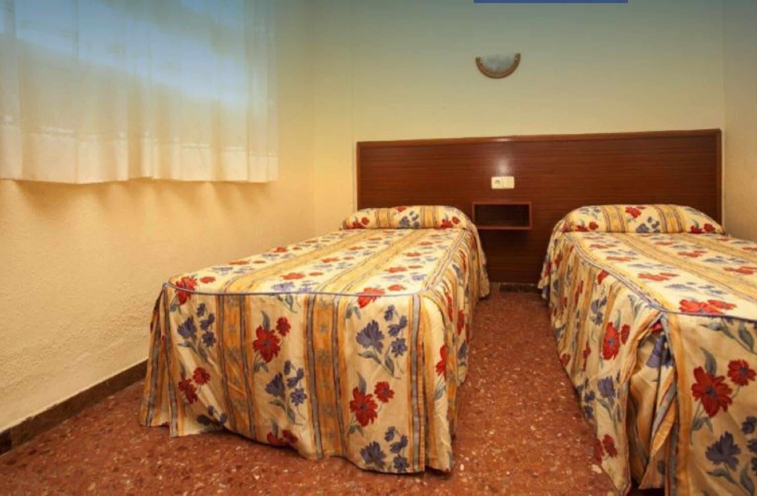 Quarto com duas camas de solteiro, cada uma com colcha de padrão floral, cabeceira de madeira e pequena prateleira entre as camas.
