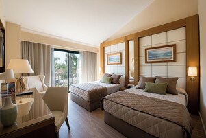 Quarto de hotel com duas camas, uma escrivaninha com abajur, uma cadeira e vista para o exterior.