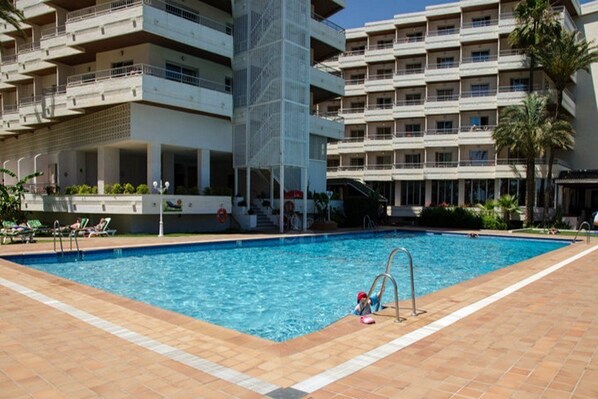 Uma piscina com deck ladrilhado, cercada por um edifício com vários andares e sacadas.