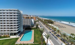 Um hotel com piscina, vista para a praia e uma estrada com carros estacionados.