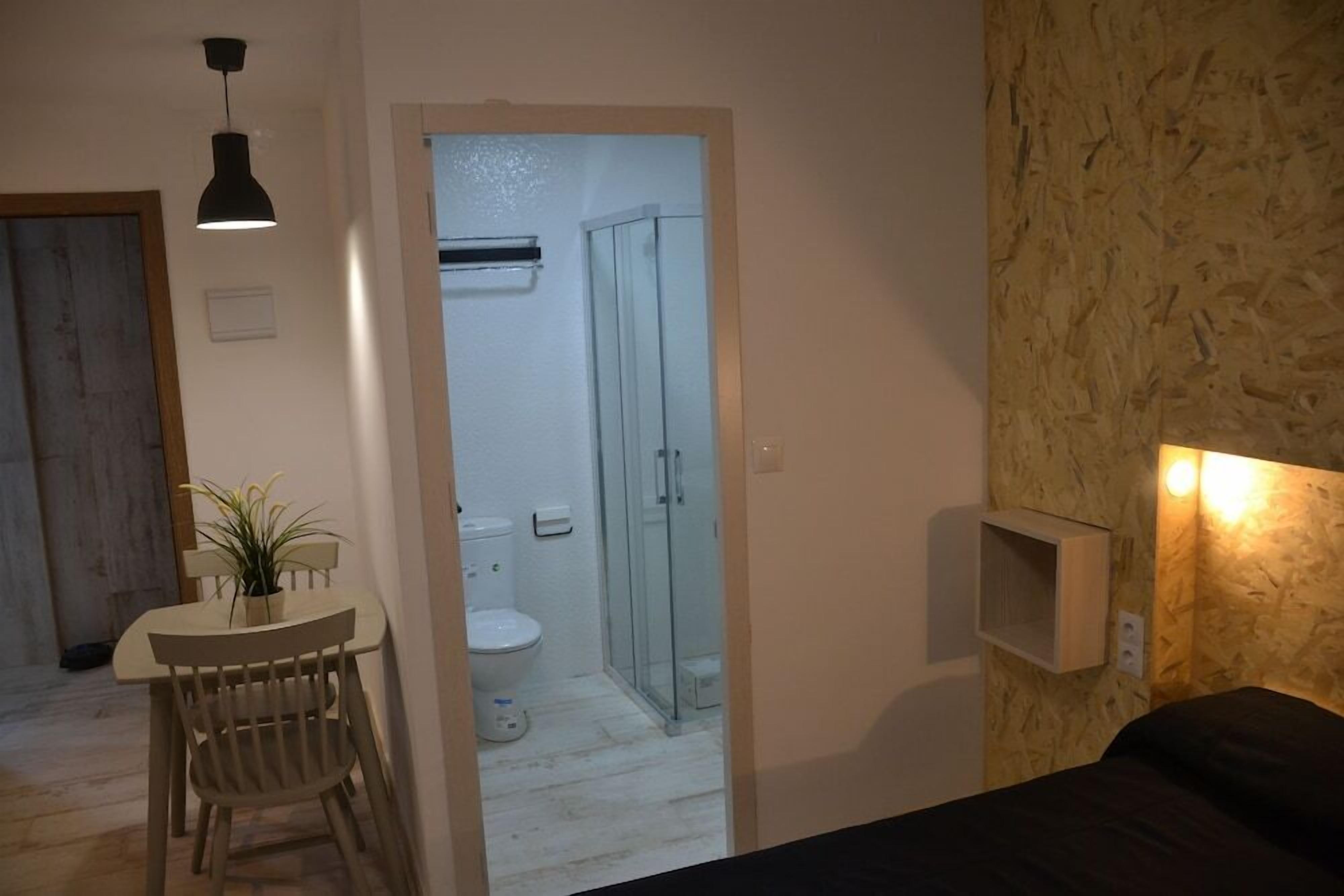 Quarto com cama, uma mesinha com cadeira, um vaso de planta e área de chuveiro visível através de uma porta aberta.