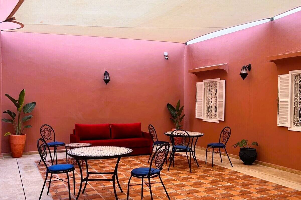 Uma área de pátio coberta com um sofá vermelho, mesas redondas e cadeiras, rodeada por plantas em vasos e painéis decorativos na parede.
