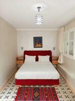 Quarto com cama, cabeceira vermelha, quadro emoldurado, mesa de cabeceira, janela com cortinas e tapete estampado.