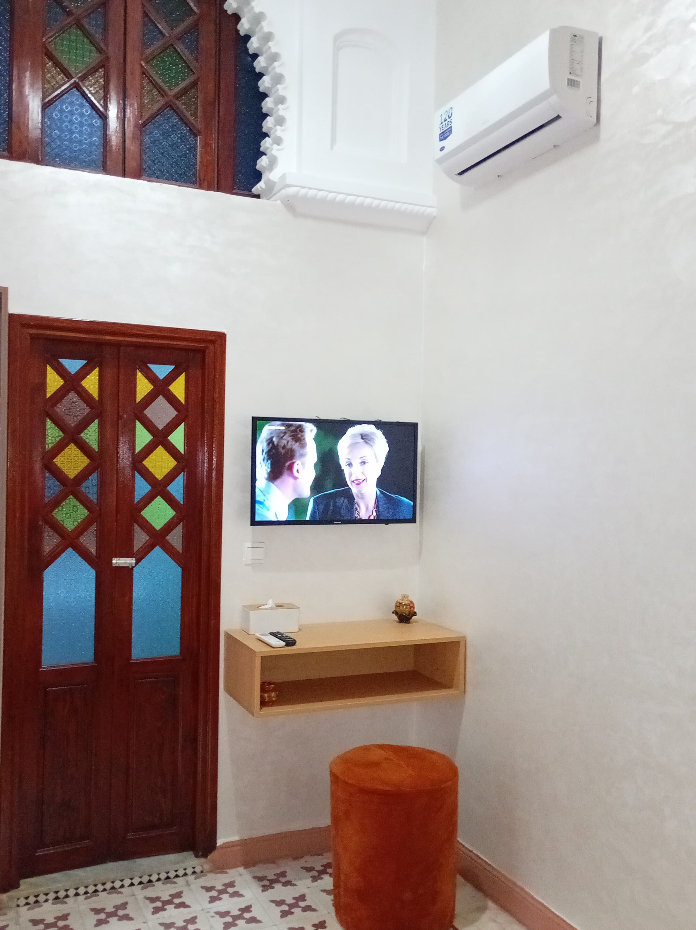 Quarto com televisão fixada na parede, porta de madeira com painéis de vidro coloridos, prateleira pequena com controle remoto e um banquinho vermelho.