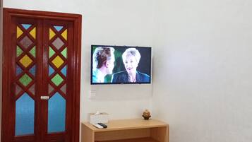 Quarto com televisão fixada na parede, porta de madeira com painéis de vidro coloridos, prateleira pequena com controle remoto e um banquinho vermelho.
