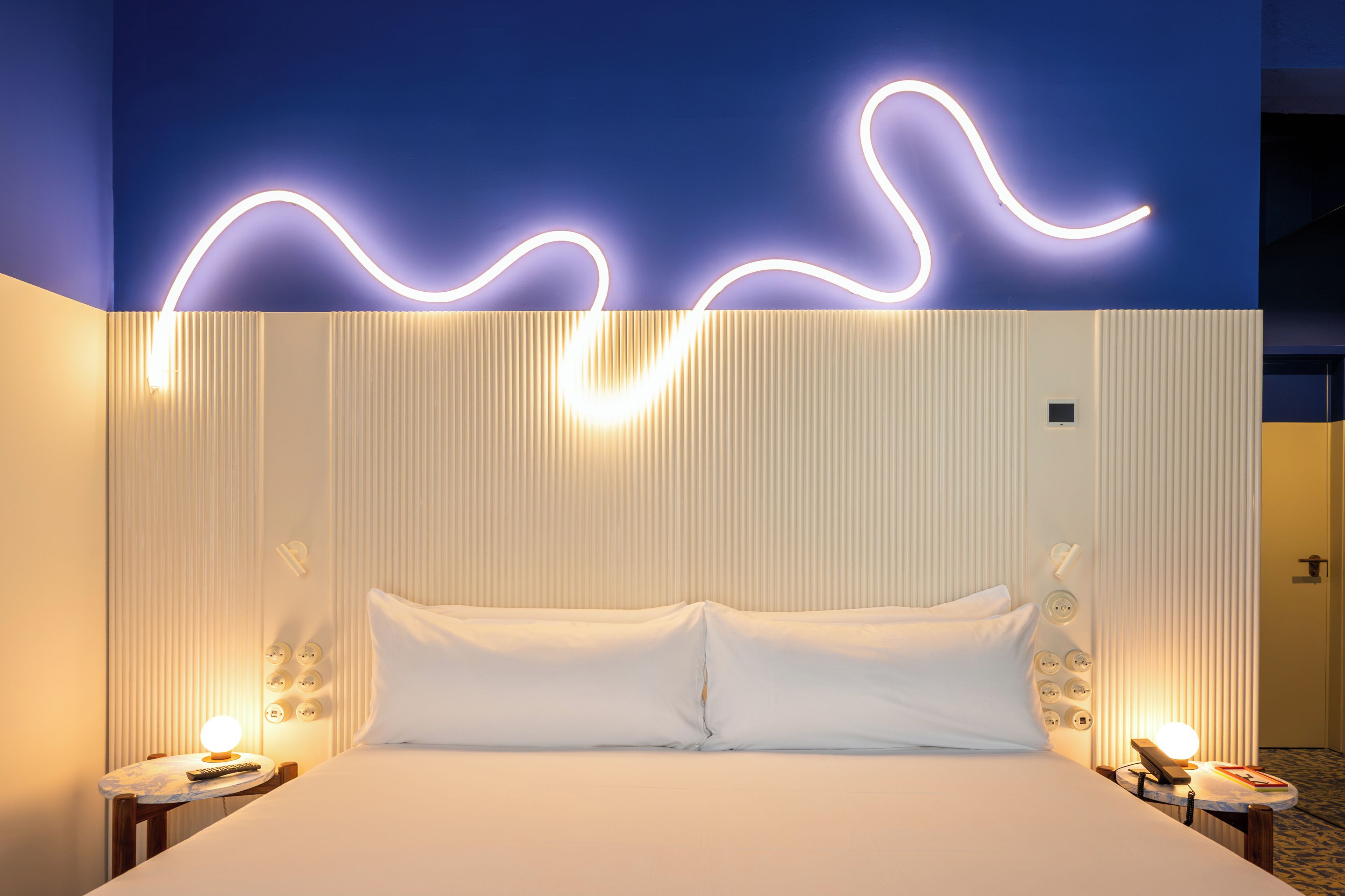 Quarto de hotel com cama, mesas de cabeceira, luminária na parede e uma porta.