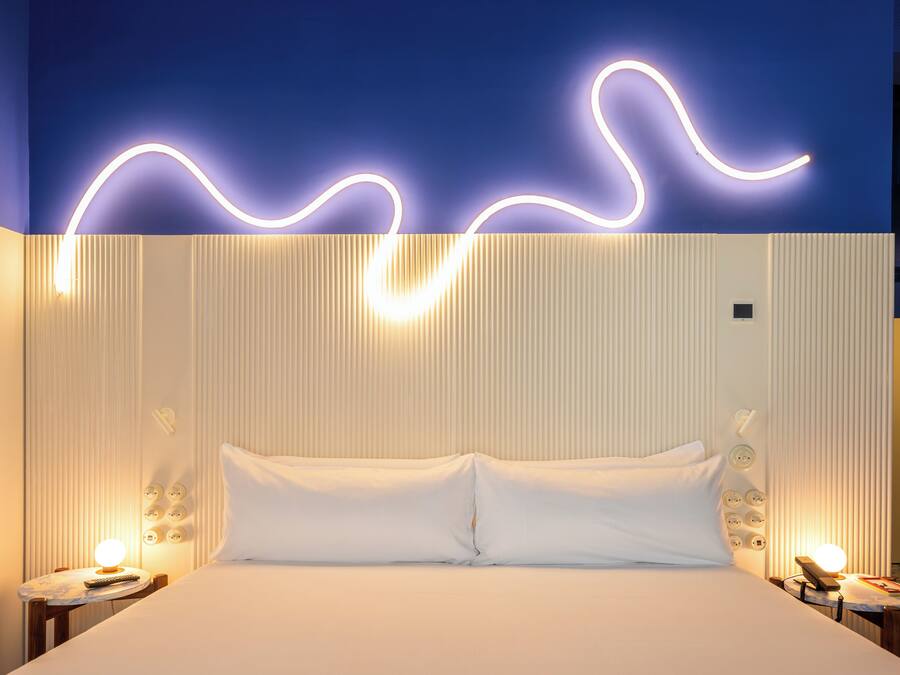 Quarto de hotel com cama, mesas de cabeceira, luminária na parede e uma porta.