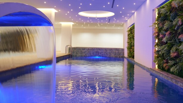 Piscina interior moderna com um recurso aquático transparente, iluminação embutida e uma parede verde com plantas.