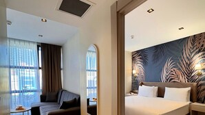 Quarto de hotel moderno com uma cama grande, um sofá, uma mesa pequena e vista para a cidade através de amplas janelas.