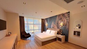 Quarto de hotel com uma cama grande, uma escrivaninha, uma cadeira, uma mesinha e uma janela ampla com cortinas.
