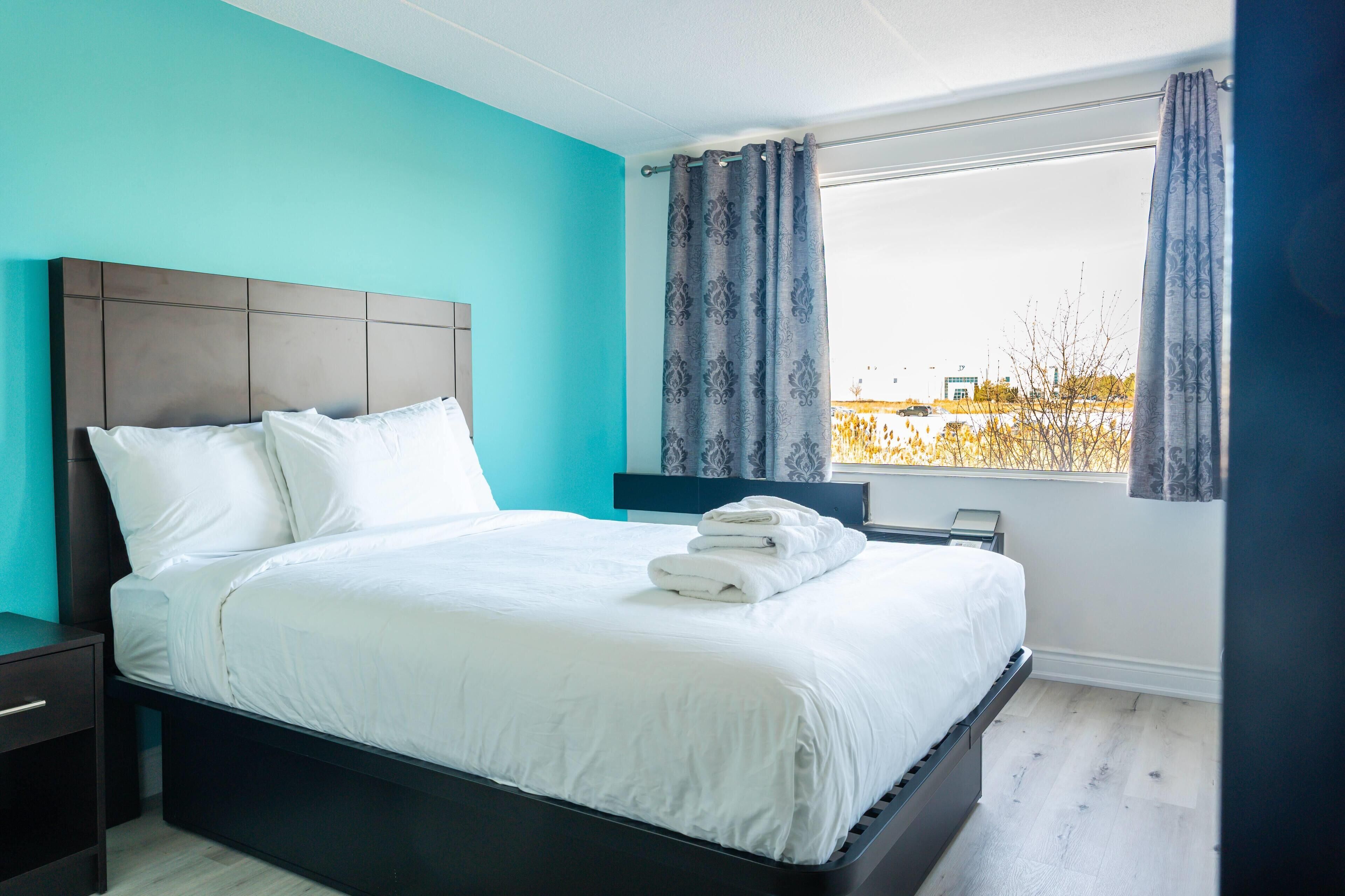 Cama bem arrumada com lençóis brancos e toalha dobrada, cabeceira escura e janela com cortinas.