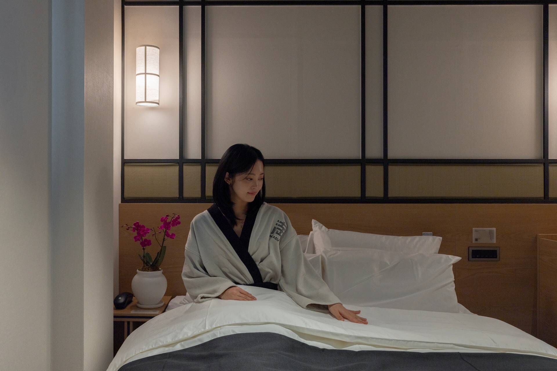 Quarto de hotel com cama, mesa de cabeceira e luminária, e um vaso com planta.