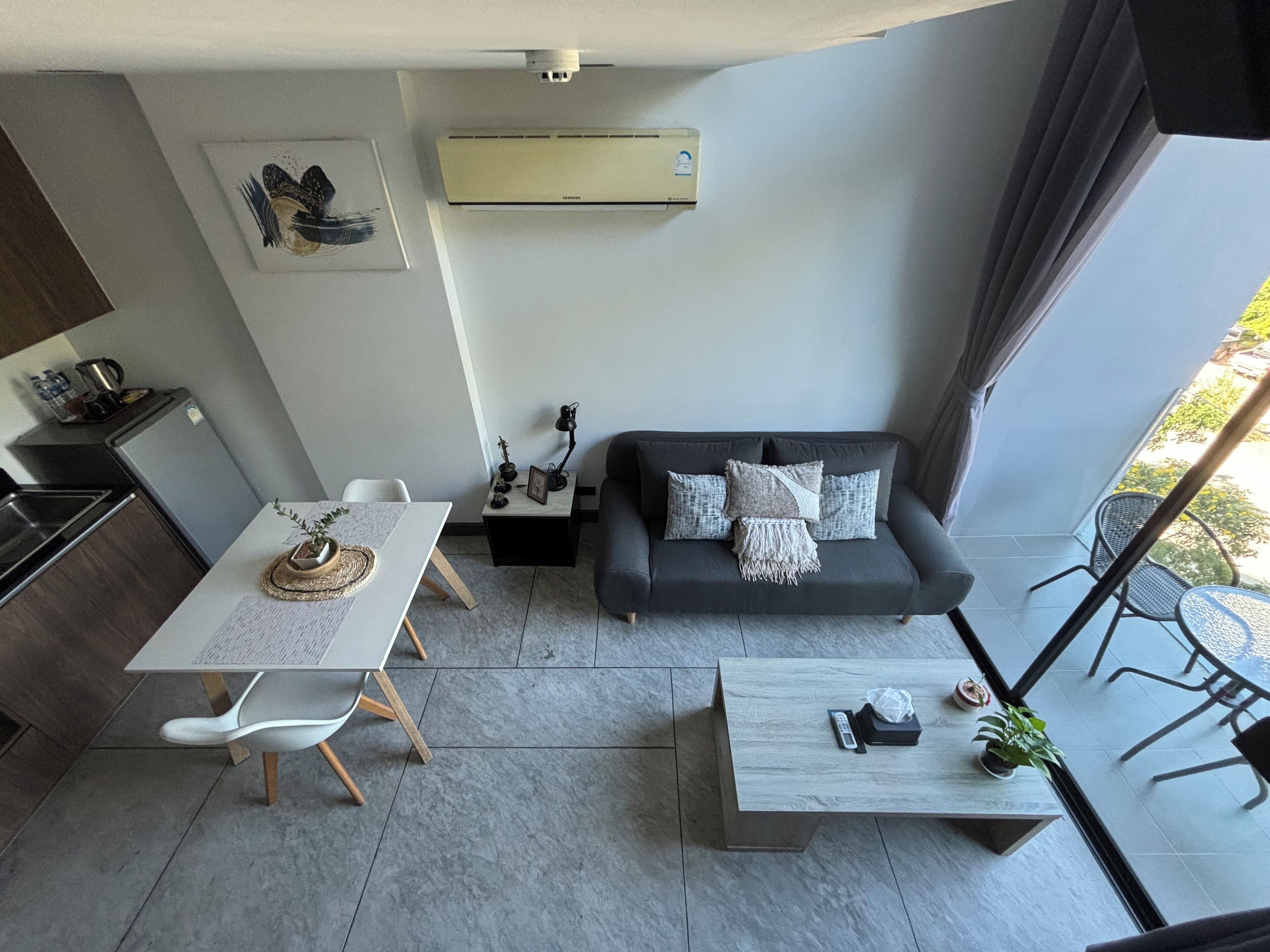 Uma sala de estar moderna com um sofá cinzento, uma mesa de jantar e uma mesa de centro. Há uma área de cozinha com frigorífico e pia. Um aparelho de ar condicionado está fixado na parede.