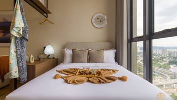 Cama arrumada com uma bandeja de petiscos e uma vela, um relógio na parede e vista da cidade através de janelas amplas.