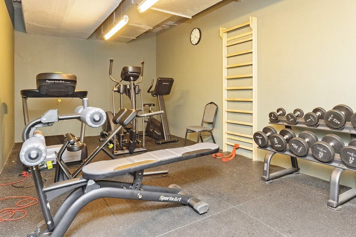 Uma academia com equipamentos de exercícios, um banco, halteres e um relógio na parede.