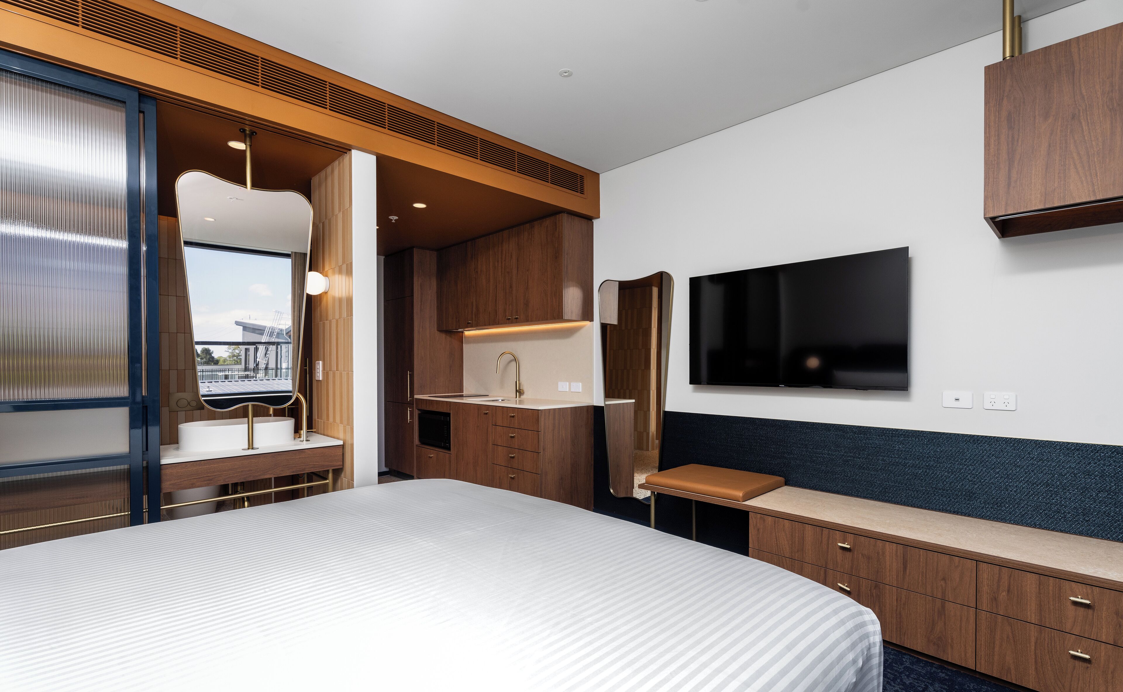 Quarto de hotel moderno com uma cama grande, banheiro com pia e espelho, e televisão fixada na parede.