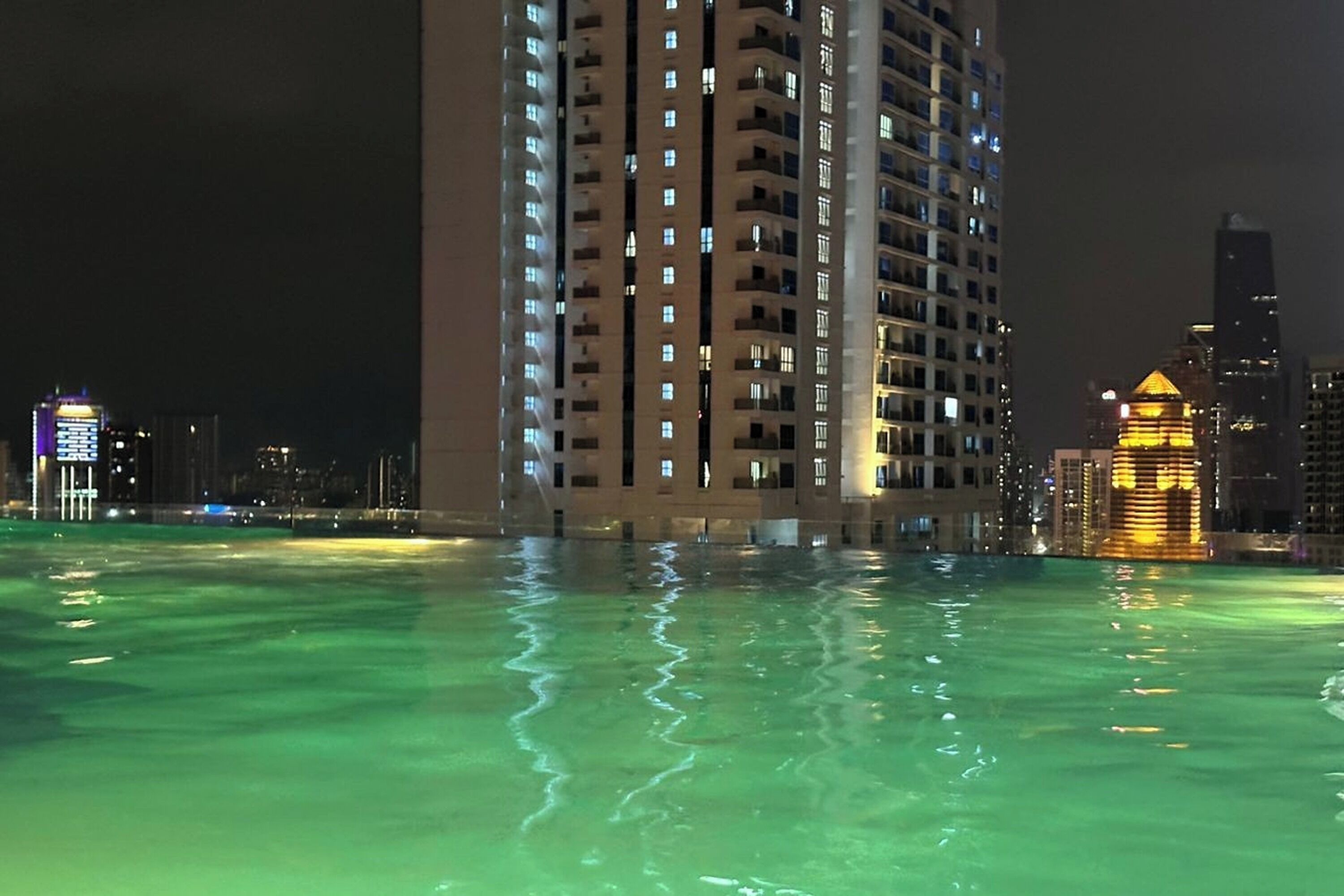 Vista noturna de um edifício alto com vários andares e varandas, refletindo em um corpo d'água.