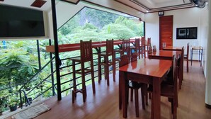 Uma sala de jantar com mesas e cadeiras de madeira, uma janela grande com vista para o verde e um ventilador de teto.