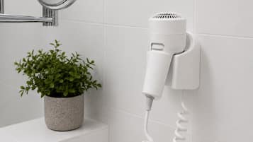 Um secador de cabelo fixado numa parede de azulejos brancos, uma planta em vaso e uma torneira de cromo.