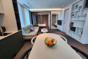Quarto de hotel moderno com sofá, mesa de jantar com uma cesta de frutas, mesinha de centro, televisão e estante de livros.