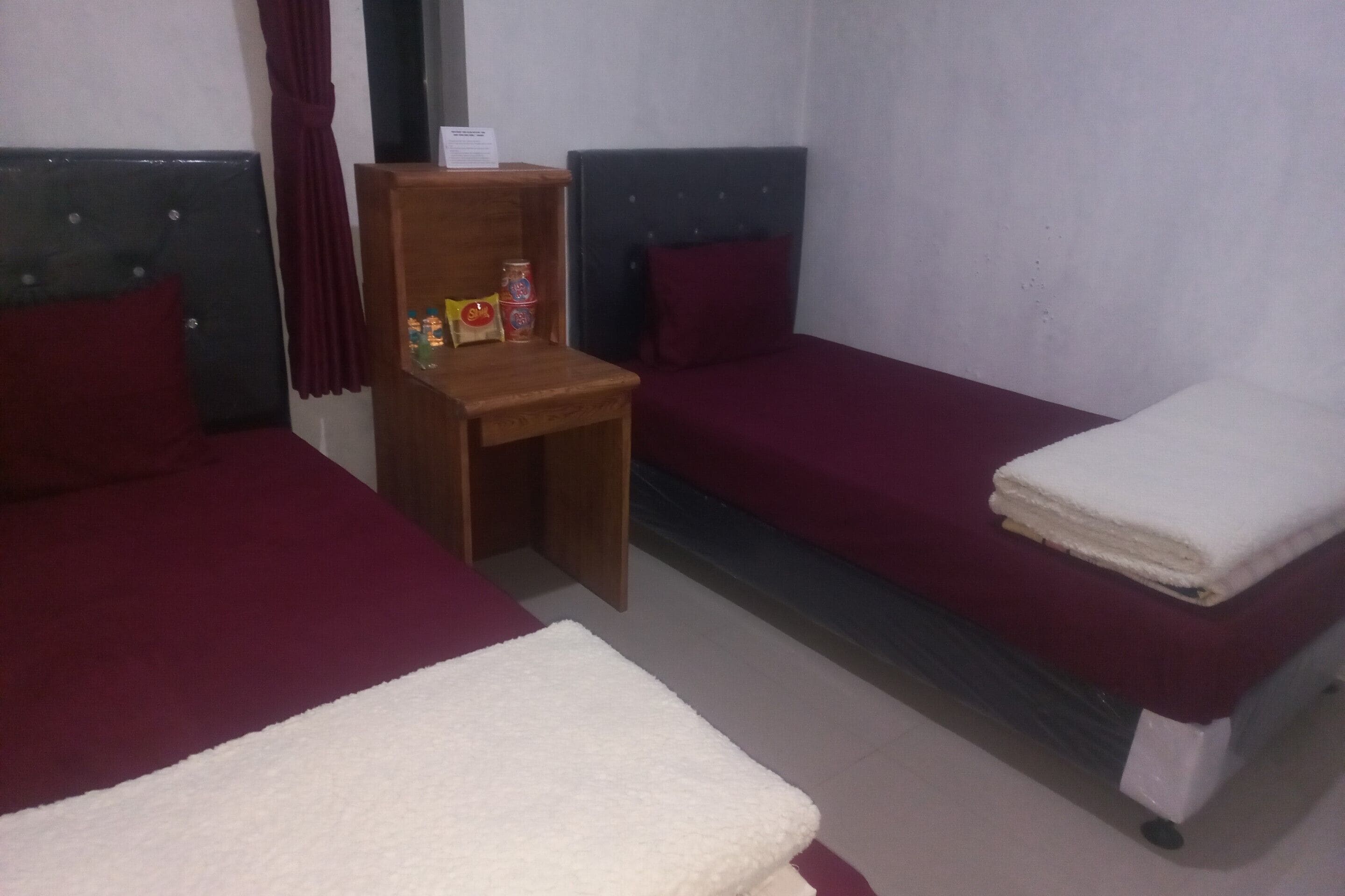 Quarto com duas camas, cada uma com cabeceira e colcha vermelha. Apresenta uma pequena mesa de cabeceira de madeira com uma lâmpada e alguns objetos.